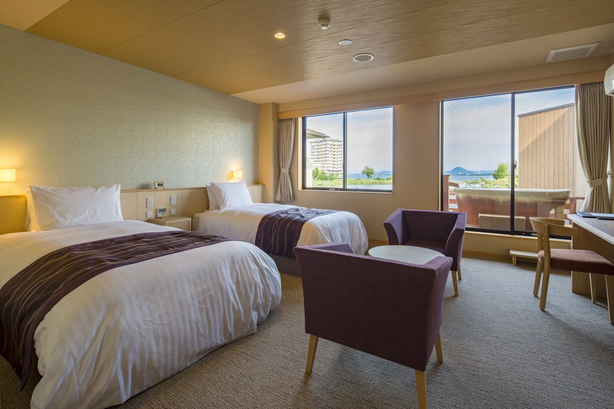 Quarto de hotel com duas camas, uma escrivaninha e uma cadeira. Há uma janela grande com vista para um edifício e áreas verdes.