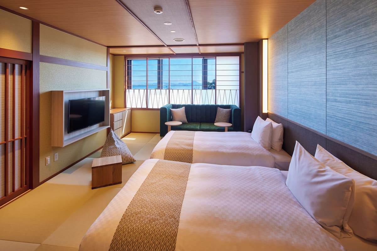 Quarto de hotel com duas camas, televisão, área de estar com vista e uma janela grande.