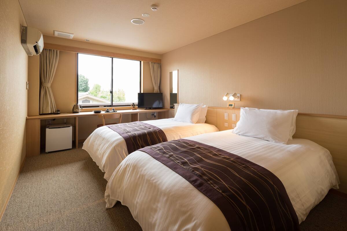 Quarto de hotel com duas camas, uma escrivaninha com televisão, um pequeno frigorífico e uma janela com cortinas.