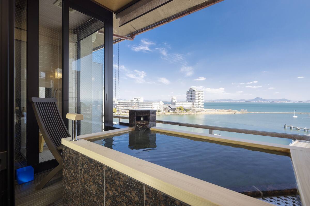 Uma casa de banho moderna com uma janela ampla, bancada de mármore e vista para o mar e edifícios.