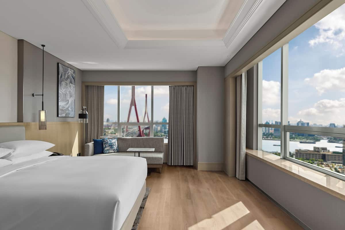 Quarto de hotel moderno com uma cama grande, área de estar e vista para a cidade através de amplas janelas.