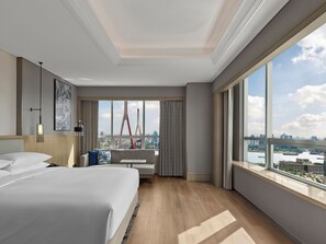Quarto de hotel moderno com uma cama grande, área de estar e vista para a cidade através de amplas janelas.