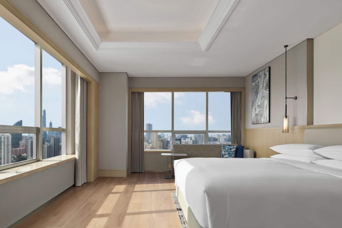 Quarto de hotel com uma grande janela, uma cama, uma escrivaninha e vista para a cidade.