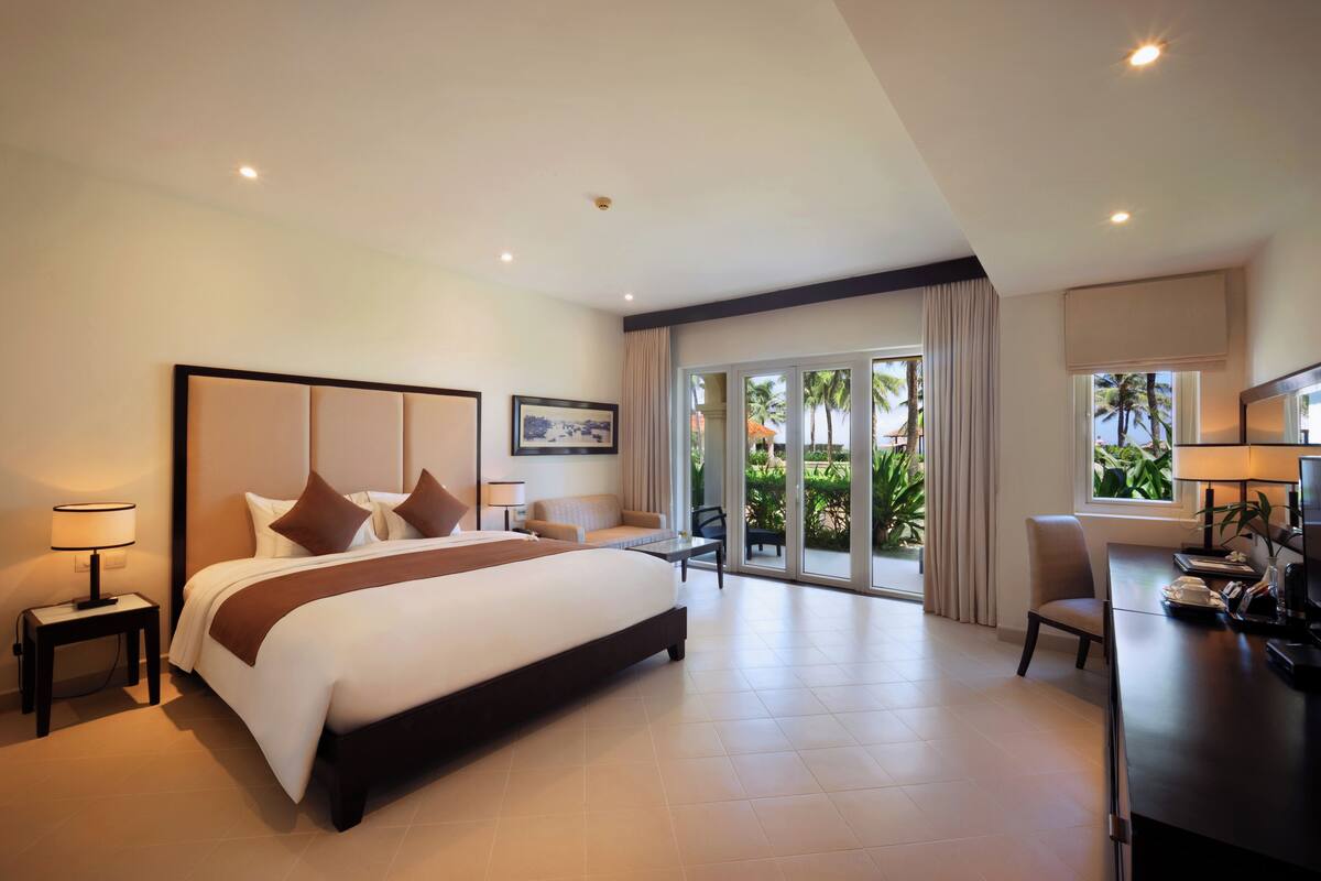 Quarto de hotel moderno com uma cama grande, uma escrivaninha e vista para palmeiras através de portas de vidro deslizantes.