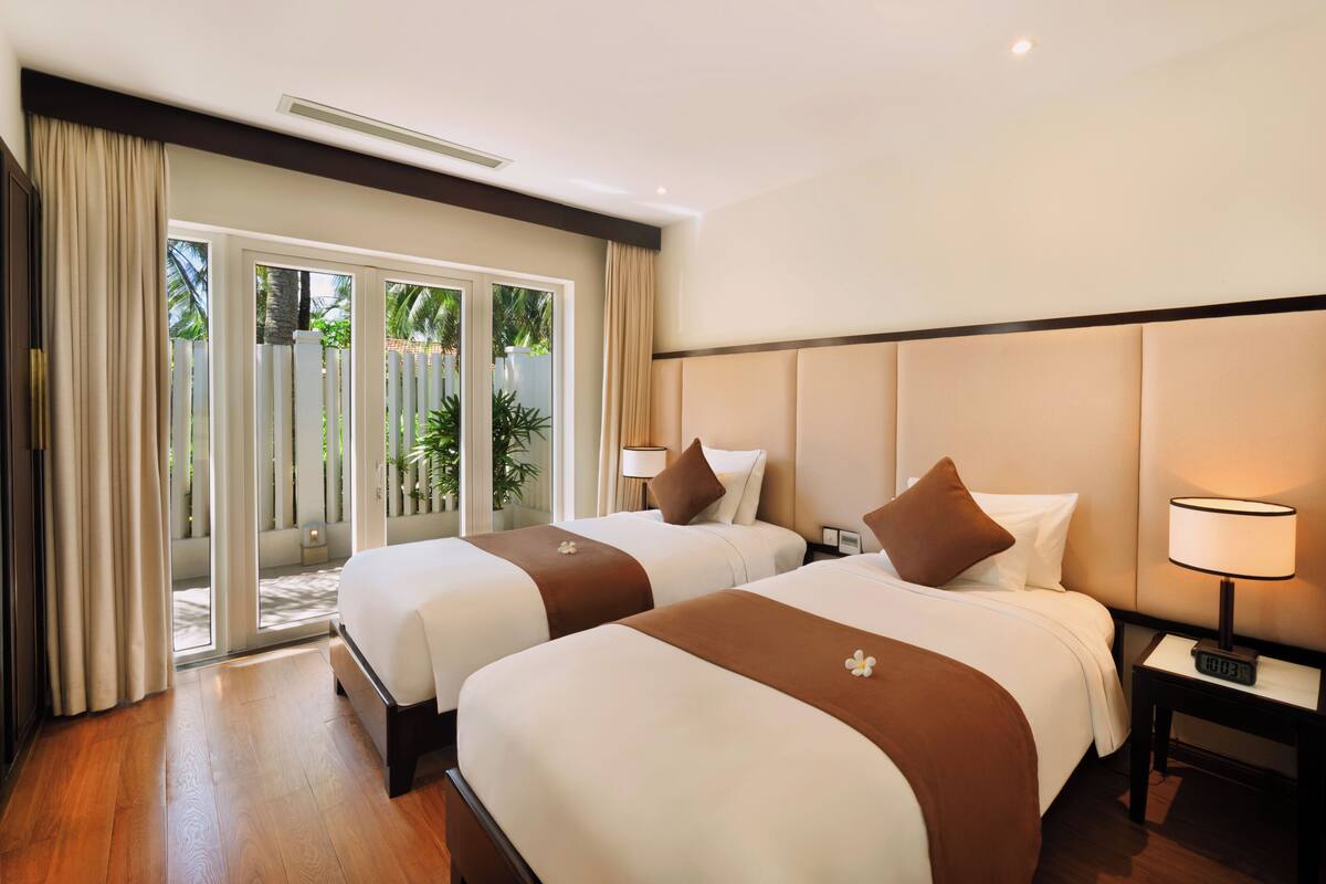 Quarto de hotel com duas camas, porta de vidro deslizante para o exterior, mesa de cabeceira com abajur e uma cabeceira grande e acolchoada.
