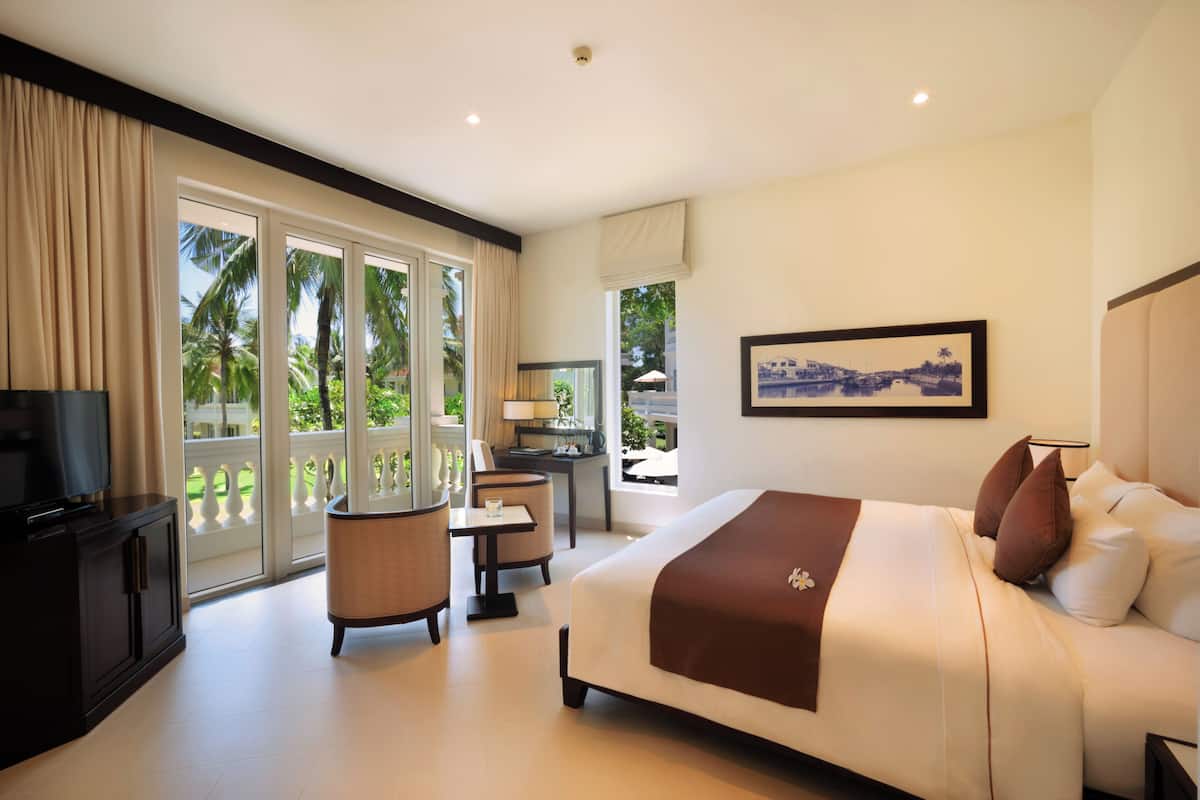 Quarto de hotel com cama, escrivaninha, cadeira, televisão e varanda com vista para palmeiras.