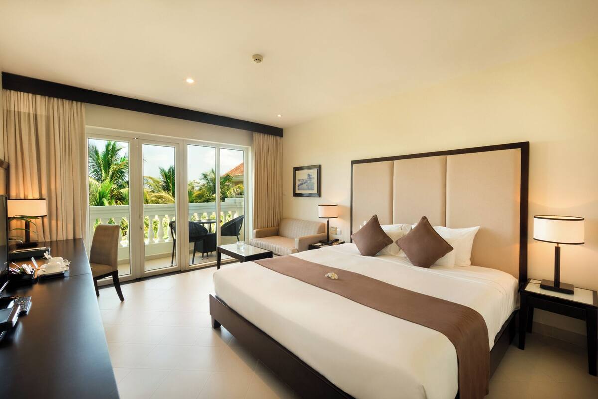 Quarto de hotel com uma cama grande, escrivaninha, cadeira, mesa pequena e varanda com vista para palmeiras.