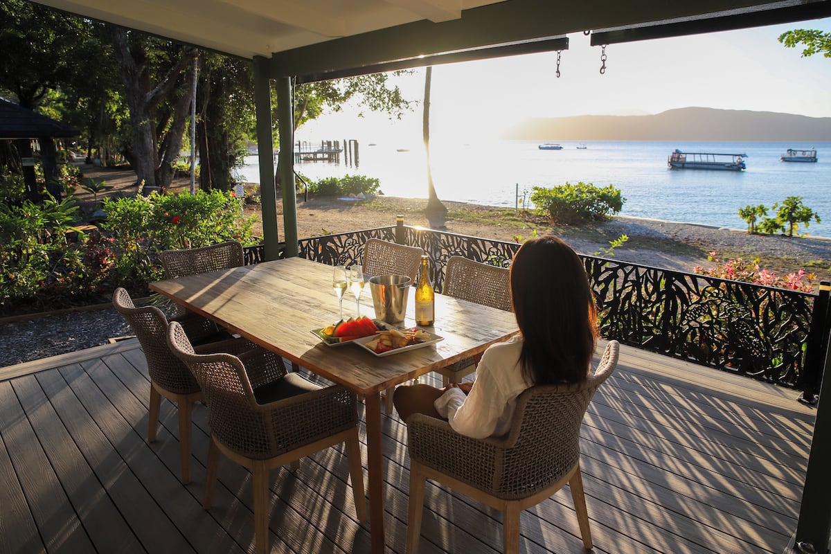 Um deck de madeira com cadeiras de vime e uma mesa, com vista para um corpo d'água calmo e barcos.