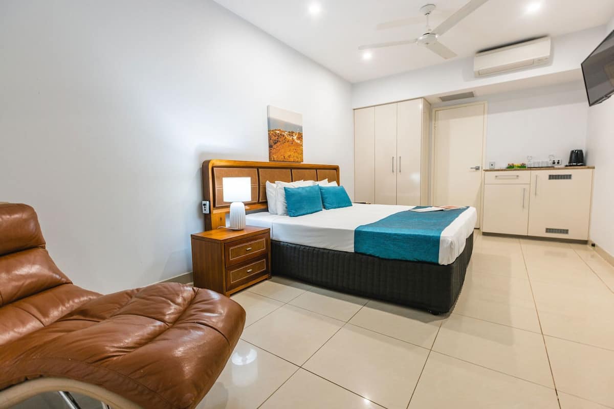 Quarto de hotel moderno com uma cama, mesa de cabeceira, poltrona de couro, guarda-roupa e cozinha compacta.