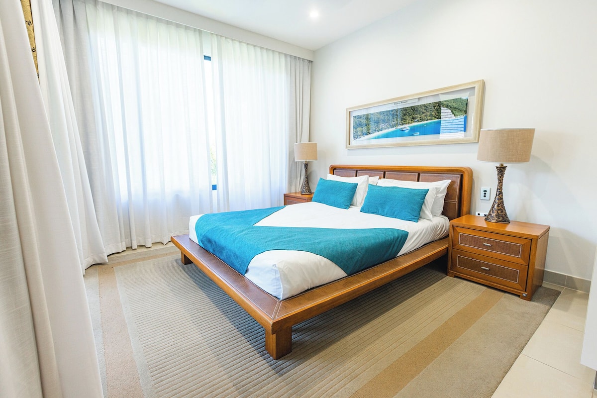 Cama bem arrumada com estrutura de madeira e roupa de cama azul, mesa de cabeceira com abajur, e uma janela grande com cortinas leves.