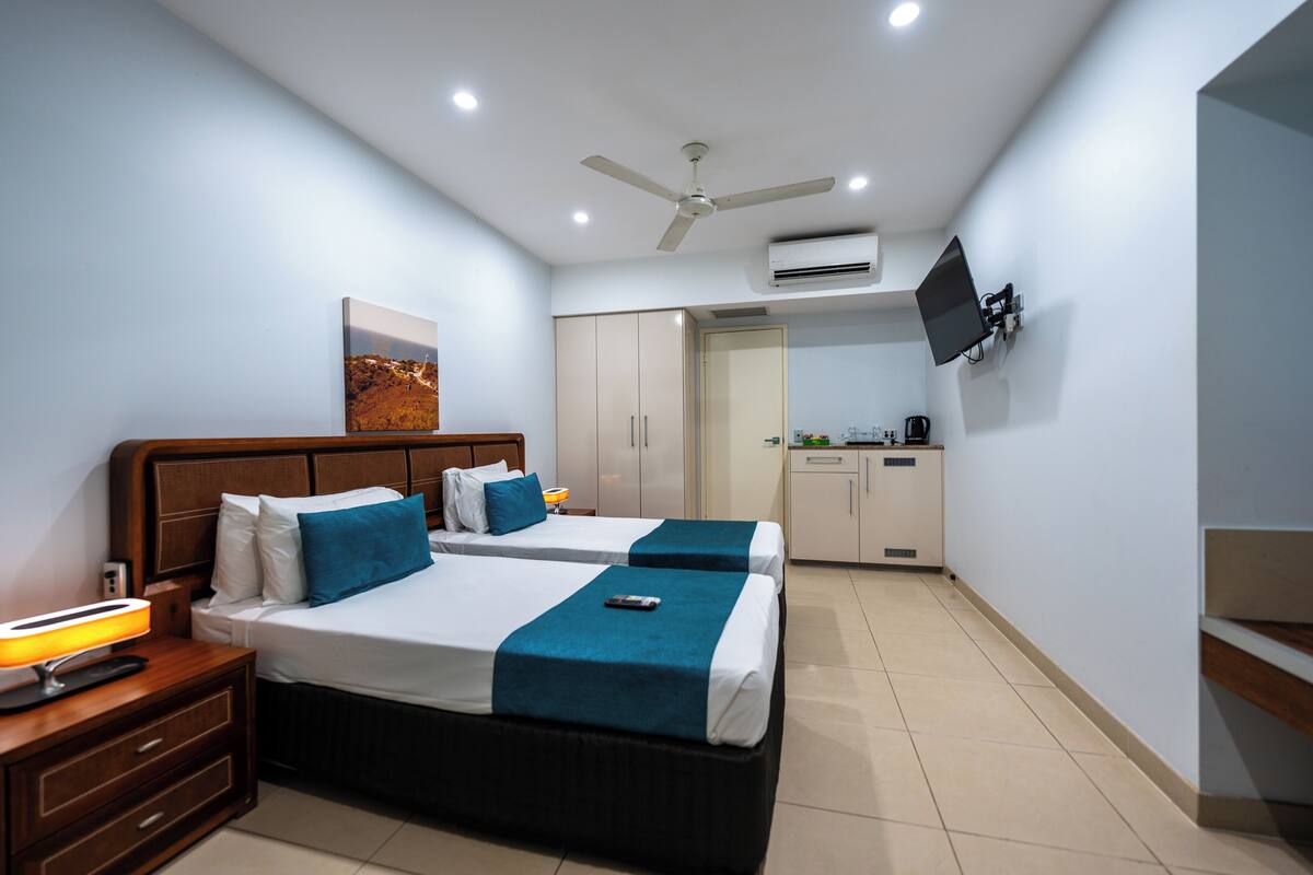 Quarto de hotel com duas camas, mesa de cabeceira, escrivaninha, ventilador de teto, ar condicionado, televisão e cozinha compacta.