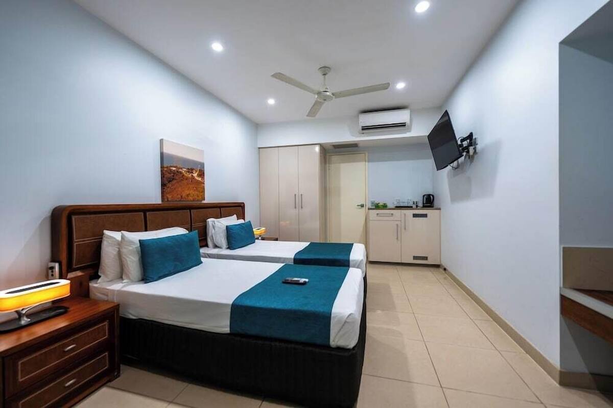 Quarto de hotel com duas camas, uma escrivaninha, uma cadeira, televisão fixada na parede e ventoinha de teto.