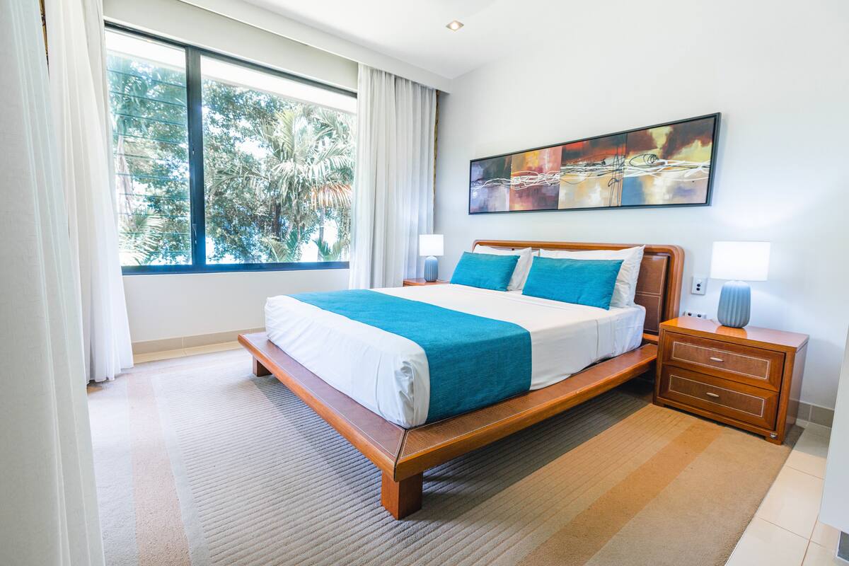 Quarto com cama grande, mesa de cabeceira, quadro na parede e janela com cortinas.