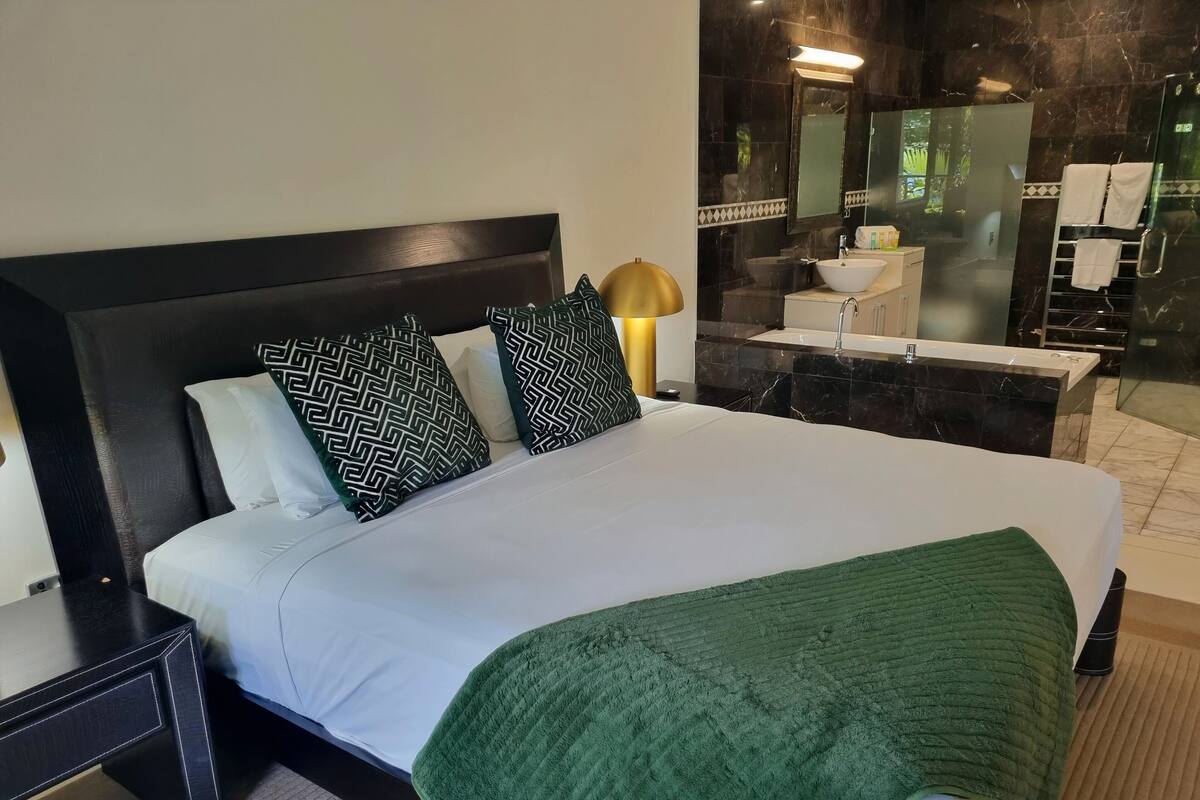 Quarto de hotel com uma cama grande, uma mesa de cabeceira, uma luminária, banheiro com pia e espelho.