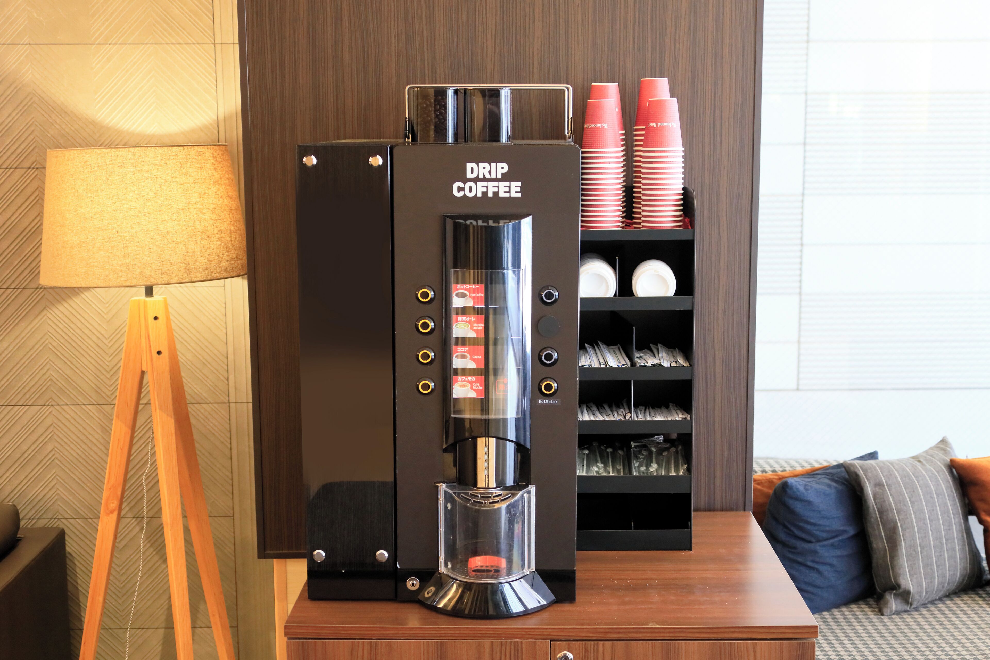 Máquina de café de filtro em um quarto moderno com uma luminária, uma prateleira com xícaras vermelhas e uma mesa lateral de madeira.