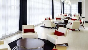 Um hall de hotel moderno com cadeiras estofadas em branco e vermelho, uma mesa redonda central e uma grande janela com cortinas translúcidas.