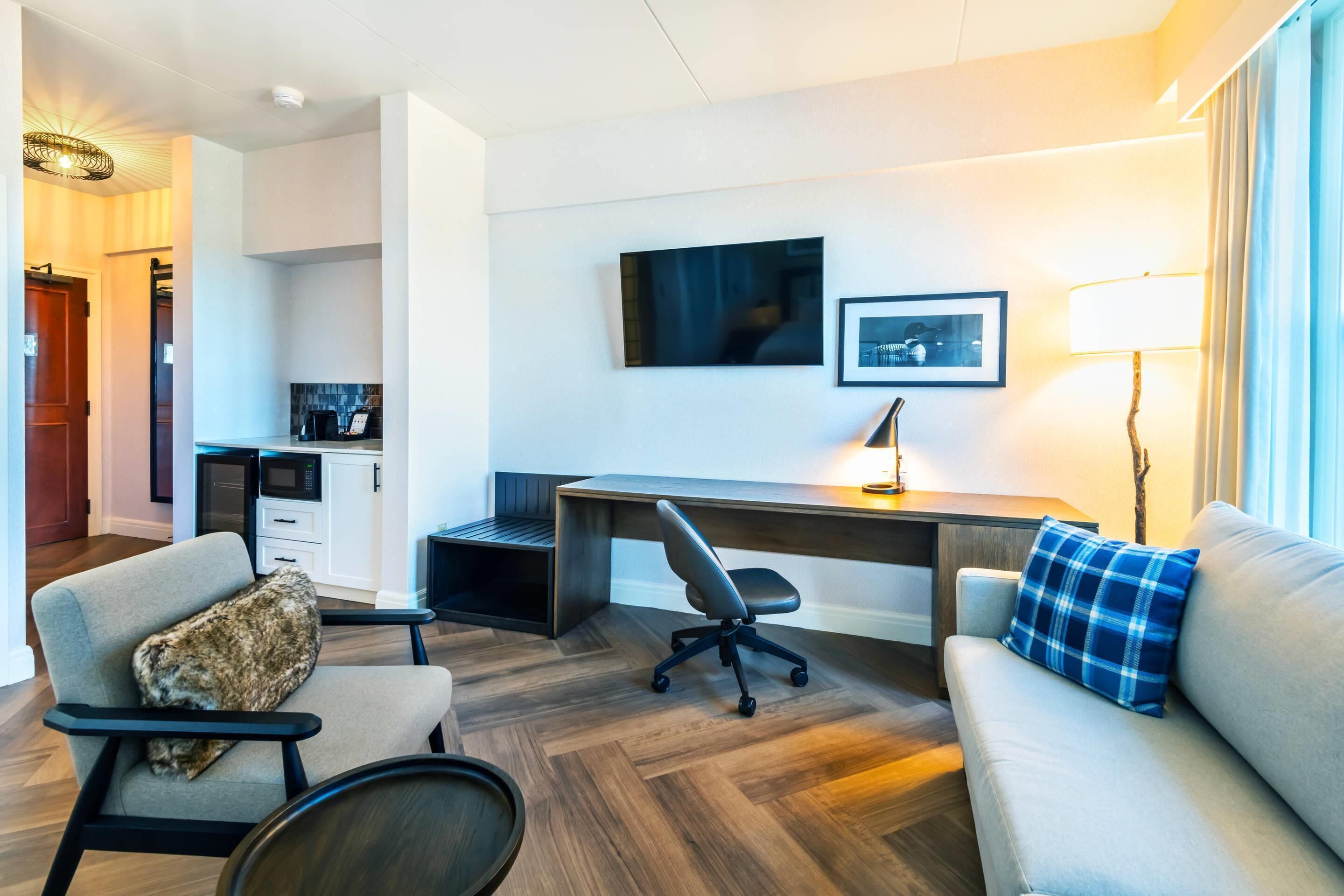 Quarto de hotel moderno com piso de madeira, um sofá, uma escrivaninha, uma cadeira, uma televisão e um abajur.