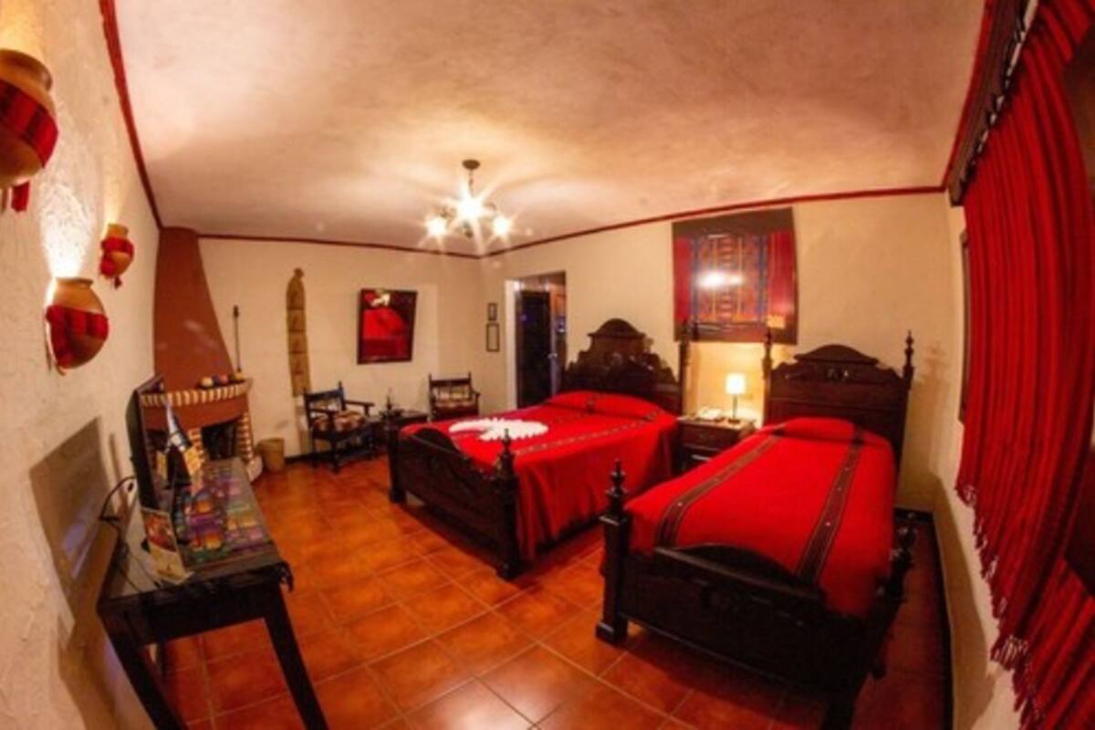 Quarto com duas camas, uma mesinha de cabeceira de madeira, uma cadeira, uma pequena mesa e um ventilador de teto.