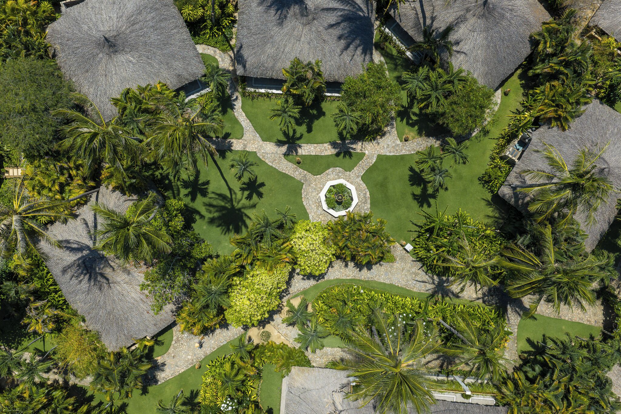 Vista aérea de um jardim paisagístico com palmeiras, um espelho d'água octogonal central e caminhos de pedra.