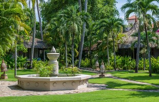 Um jardim tropical com uma fonte central, palmeiras e um edifício com telhado de palha.