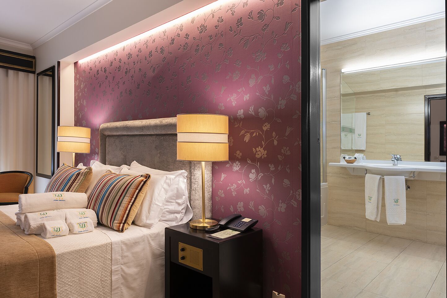 Quarto de hotel com cama, duas luminárias de cabeceira, mesa de cabeceira e banheiro com pia e espelho.