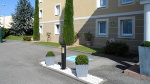 Um edifício de vários andares com fachada bege, janelas retangulares e estacionamento com brita branca e plantas em vasos.