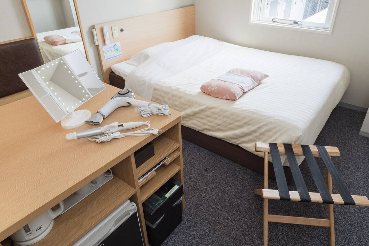Quarto de hotel com cama, escrivaninha com espelho, secador de cabelo e um banquinho.