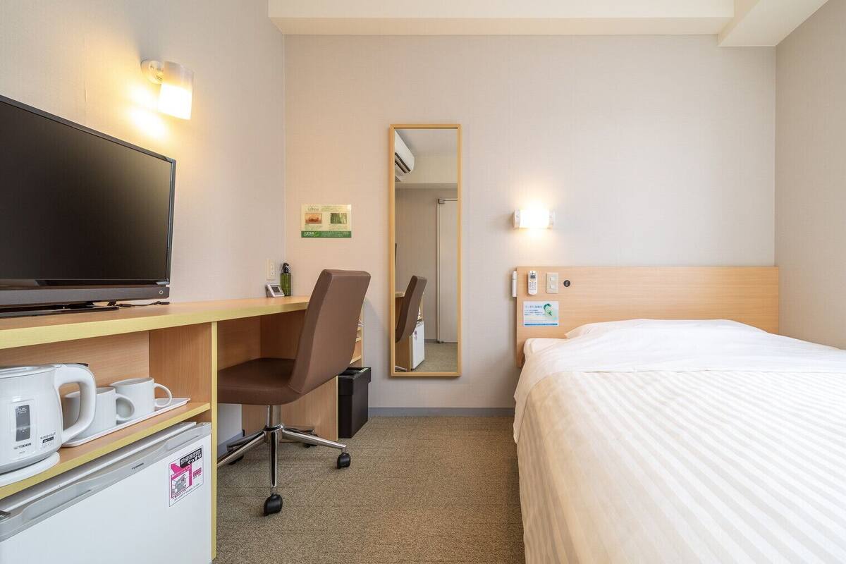 Quarto de hotel com cama, escrivaninha, cadeira, televisão e espelho.