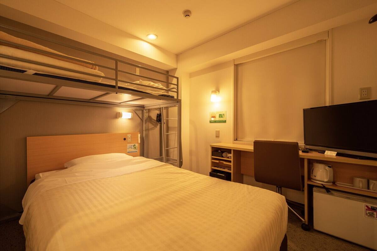 Quarto de hotel com beliche, uma escrivaninha com televisão e um frigorífico pequeno.