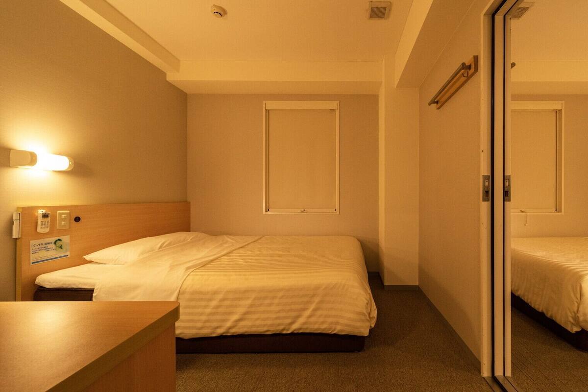 Quarto de hotel com duas camas, uma mesa de cabeceira, uma luminária na parede e uma porta com espelho.