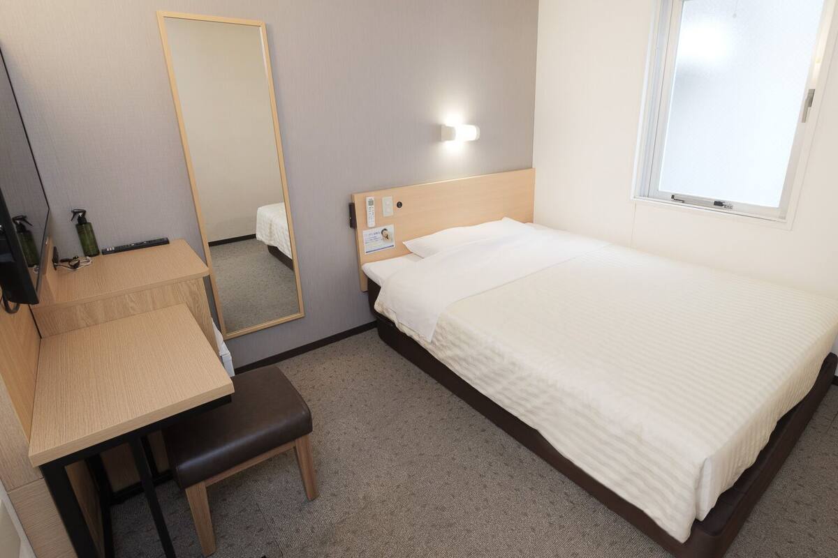 Quarto de hotel com cama, escrivaninha com cadeira, espelho, mesa de cabeceira, luminária na parede e janela.