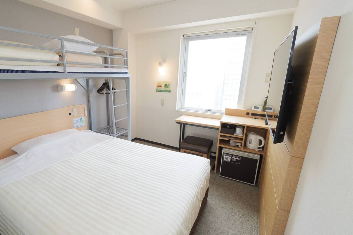 Quarto de hotel com beliche, uma mesa com cadeira, televisão de tela plana e uma pequena cozinha.