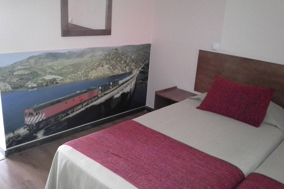 Quarto de hotel com cama, mesa de cabeceira, travesseiro vermelho e um mural na parede retratando um trem sobre uma ponte.