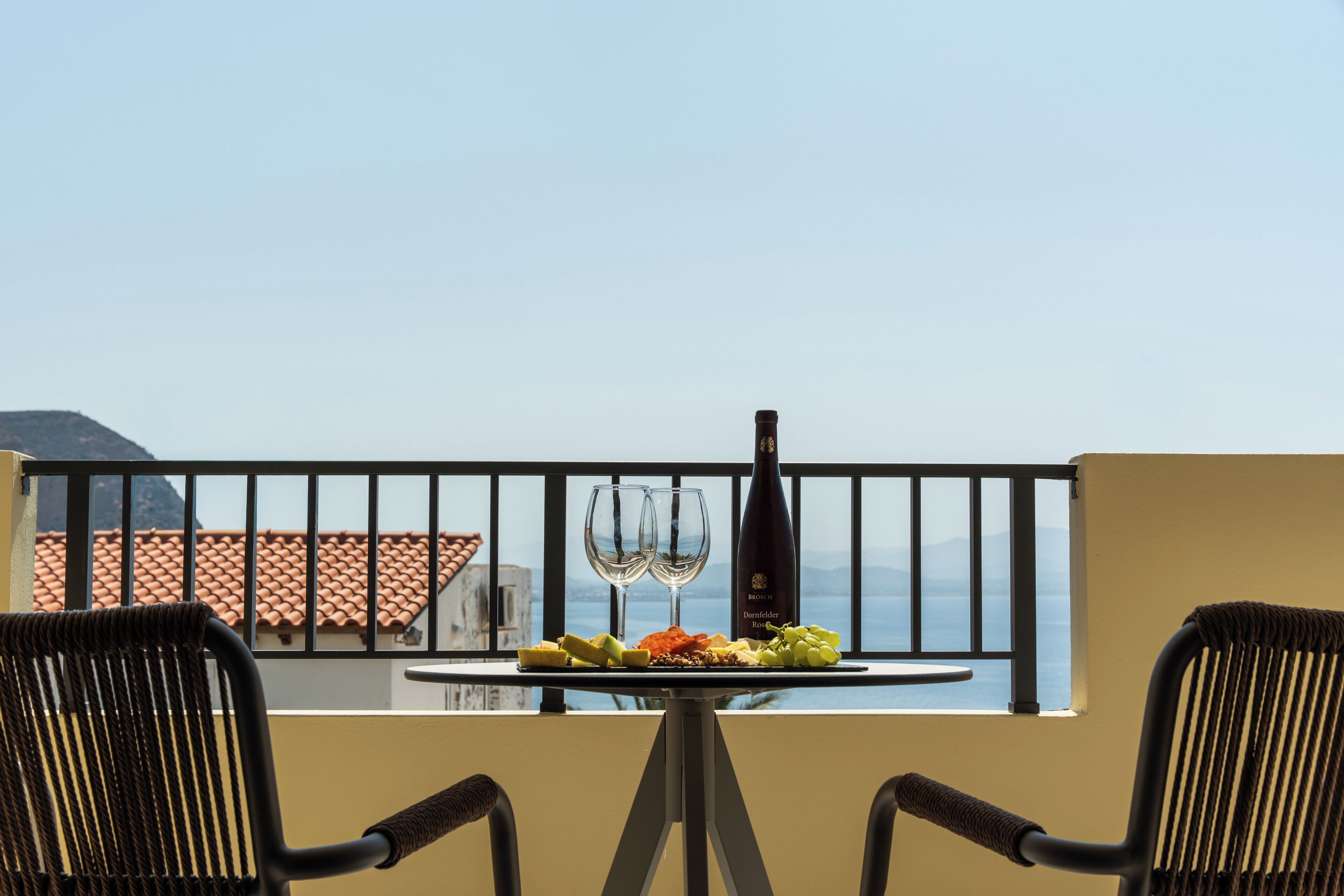 Uma mesa com uma garrafa de vinho, dois copos e um prato de fruta numa varanda com vista para o mar e montanhas.