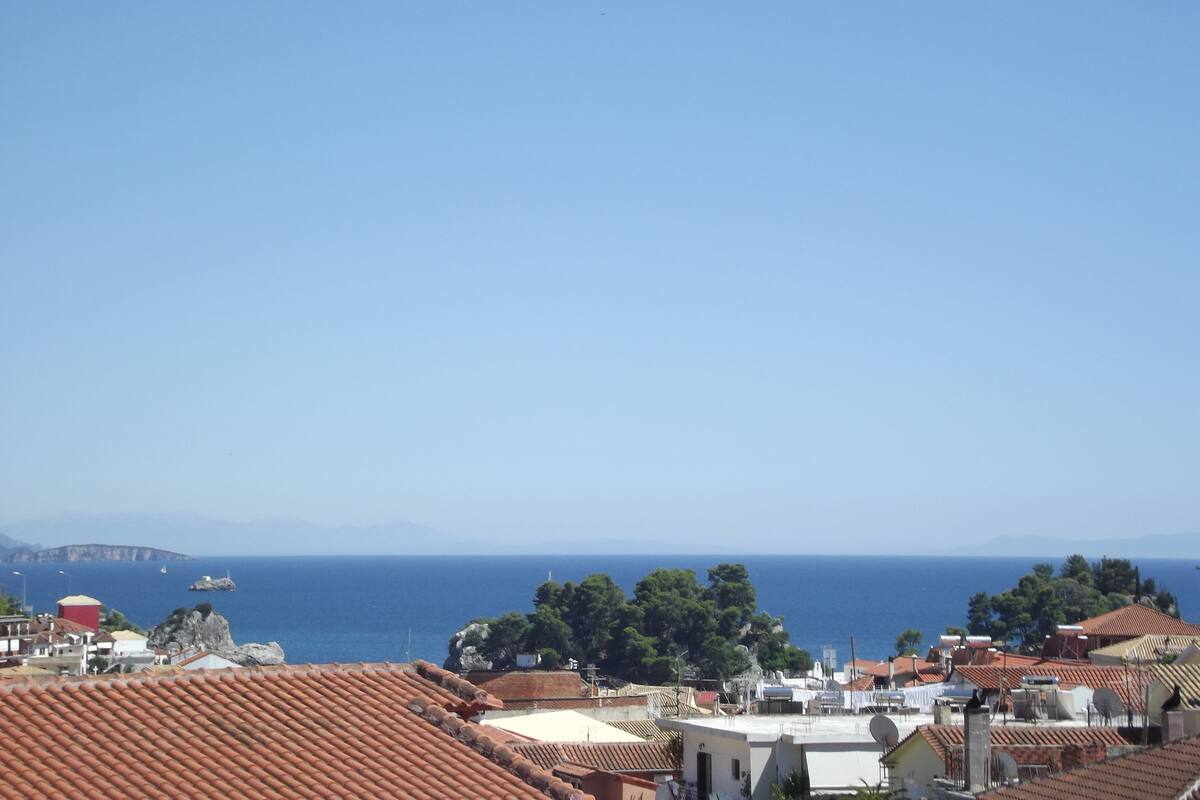 Uma vila costeira com telhados de terracota, céu azul claro e o mar ao fundo.