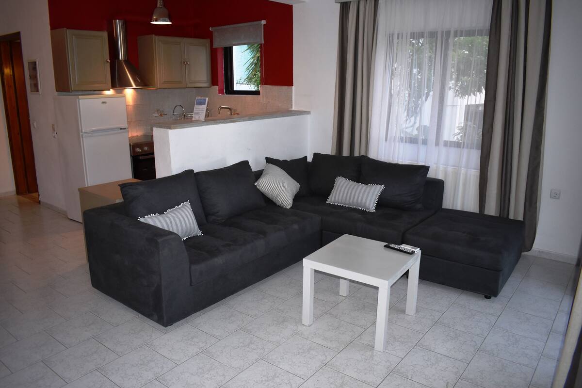 Uma sala de estar moderna com sofá seccional, mesa de centro branca e área de cozinha com geladeira, fogão e pia.