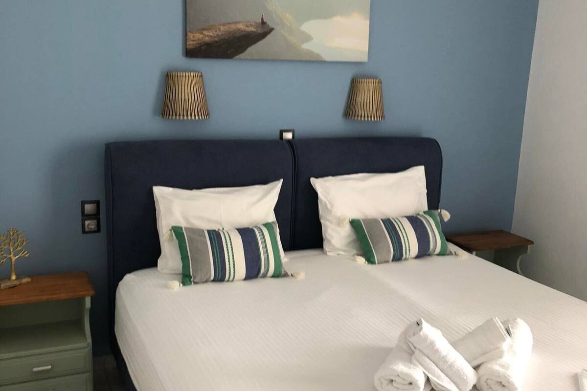 Quarto de hotel com uma cama grande, duas luminárias de cabeceira, um quadro na parede e uma mesinha de cabeceira de madeira.