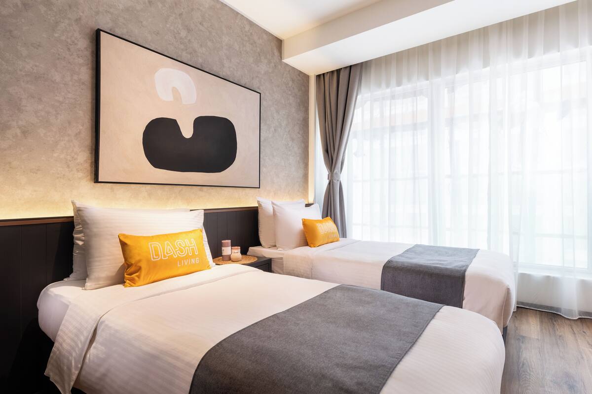 Quarto de hotel com duas camas, uma grande obra de arte abstrata na parede e vista para o exterior através de cortinas translúcidas.