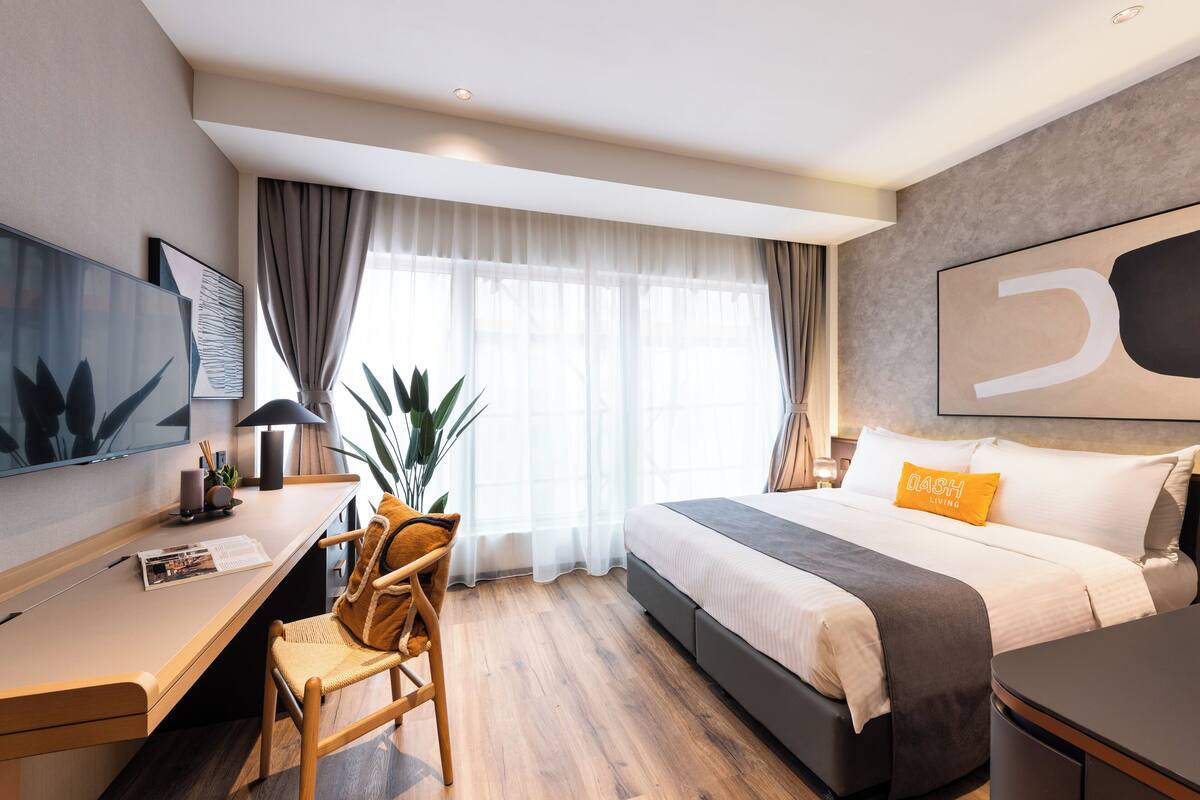 Quarto de hotel moderno com cama, escrivaninha, cadeira e uma grande janela com cortinas.
