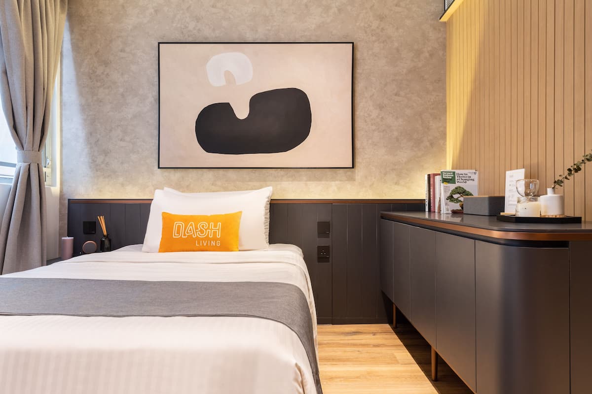 Quarto de hotel moderno com uma cama bem arrumada, mesa de cabeceira com livros e um abajur, e um quadro abstrato grande na parede.