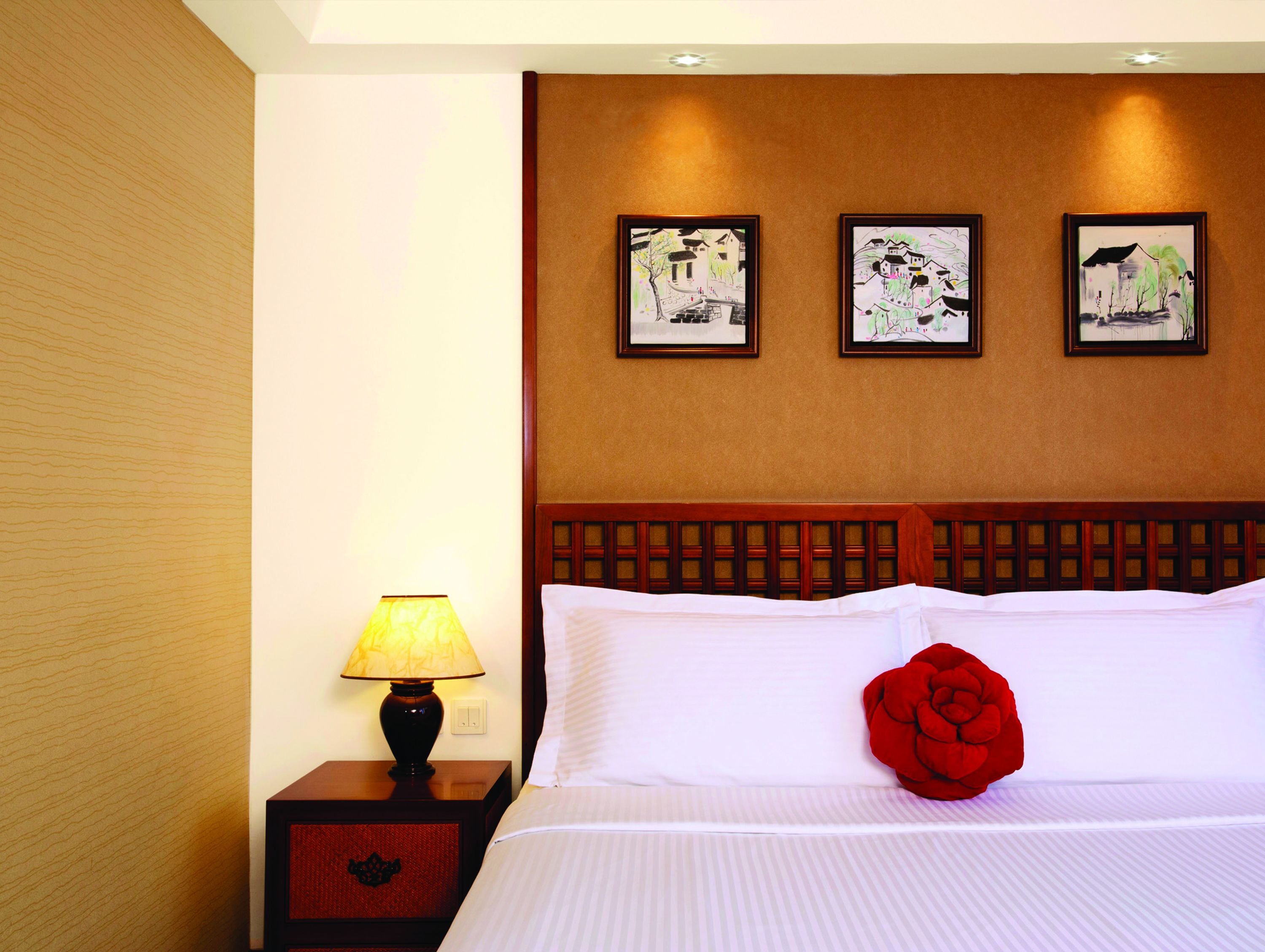 Quarto de hotel com cabeceira de madeira, cama com roupa de cama branca e uma decoração com uma rosa vermelha, mesa de cabeceira com abajur e três obras de arte emolduradas na parede.