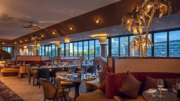 Um restaurante moderno com mesas redondas, assentos confortáveis e grandes janelas com vista para a cidade.