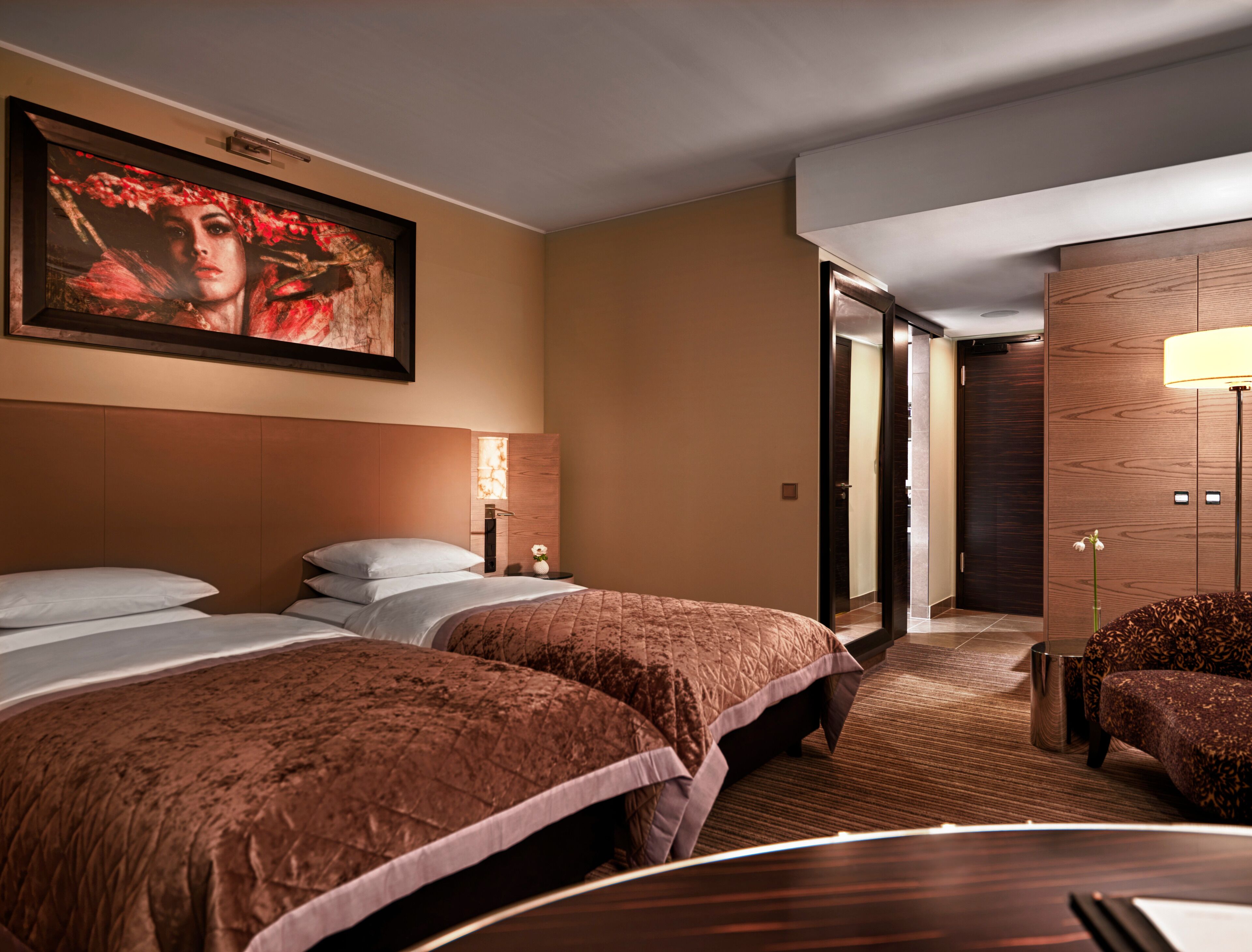 Quarto de hotel com duas camas, um quadro grande, uma luminária, um espelho e um armário de madeira.