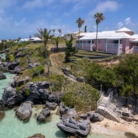 Uma propriedade à beira-mar com um edifício rosa, costa rochosa, água turquesa cristalina e palmeiras.