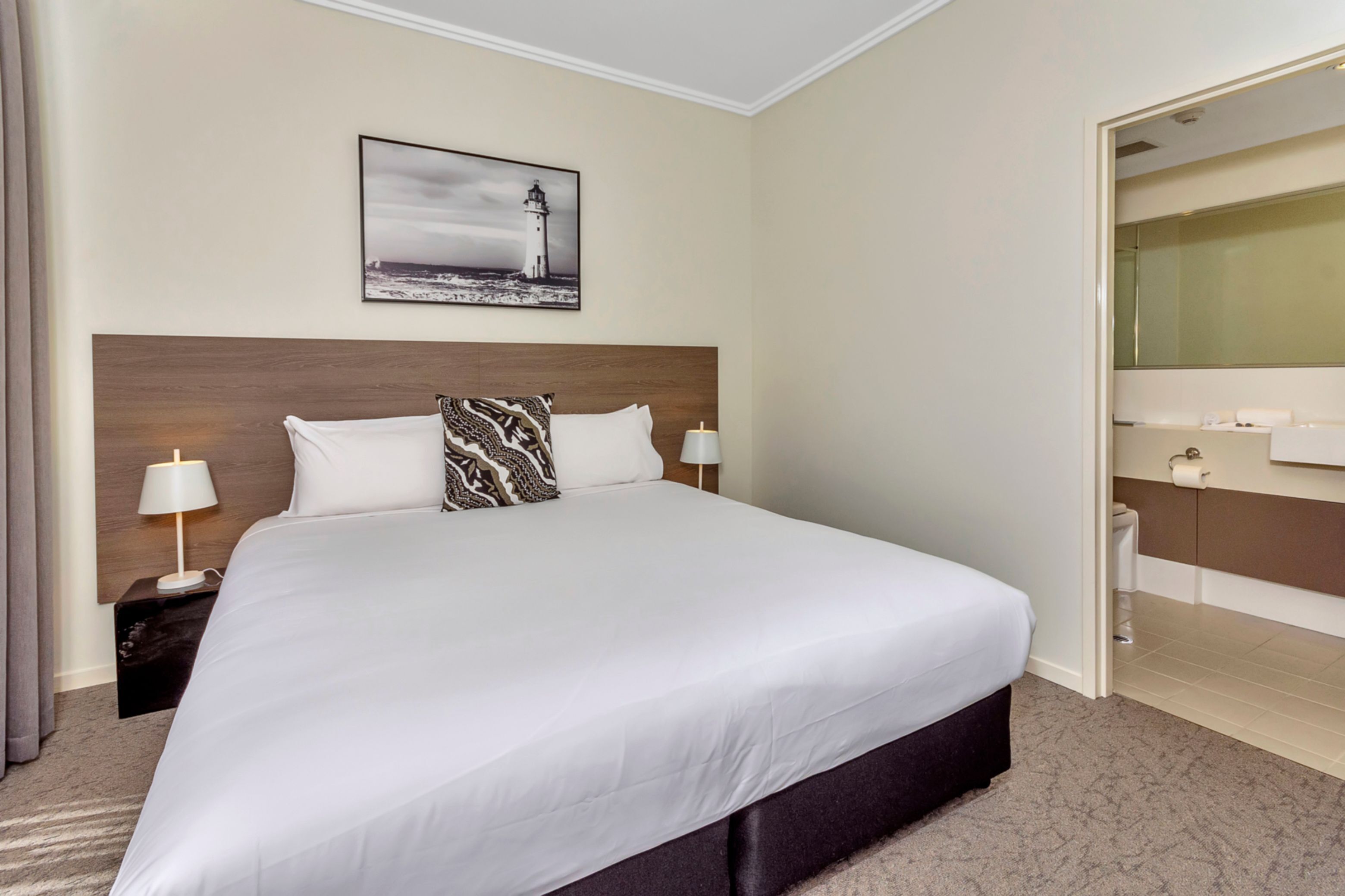 Quarto de hotel com uma cama grande, cabeceira, mesa de cabeceira com luminária, banheiro com espelho e um quadro na parede.