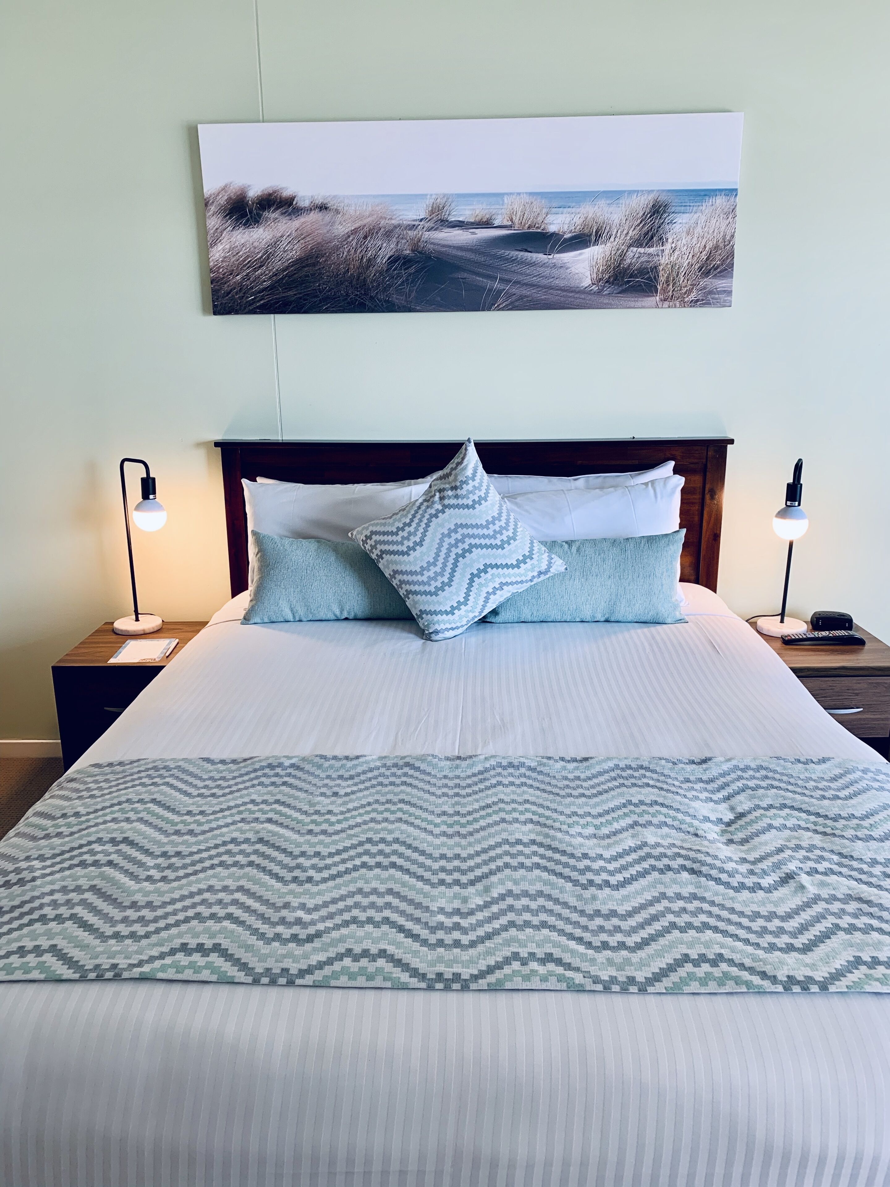 Uma cama bem arrumada com colcha estampada e almofadas, com mesas de cabeceira laterais com abajures, em um quarto com um grande quadro de paisagem na parede.