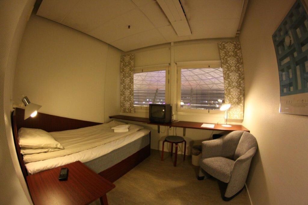 Quarto de hotel pequeno e compacto, com uma cama de solteiro, escrivaninha com televisão, cadeira e janela com vista para a cidade.