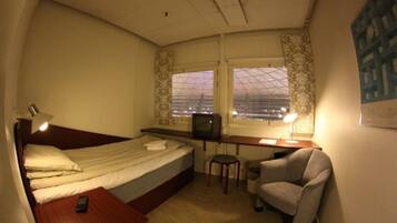 Quarto de hotel pequeno e compacto, com uma cama de solteiro, escrivaninha com televisão, cadeira e janela com vista para a cidade.