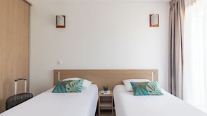 Quarto de hotel com duas camas de solteiro, uma mesinha com uma planta e uma mala no chão.