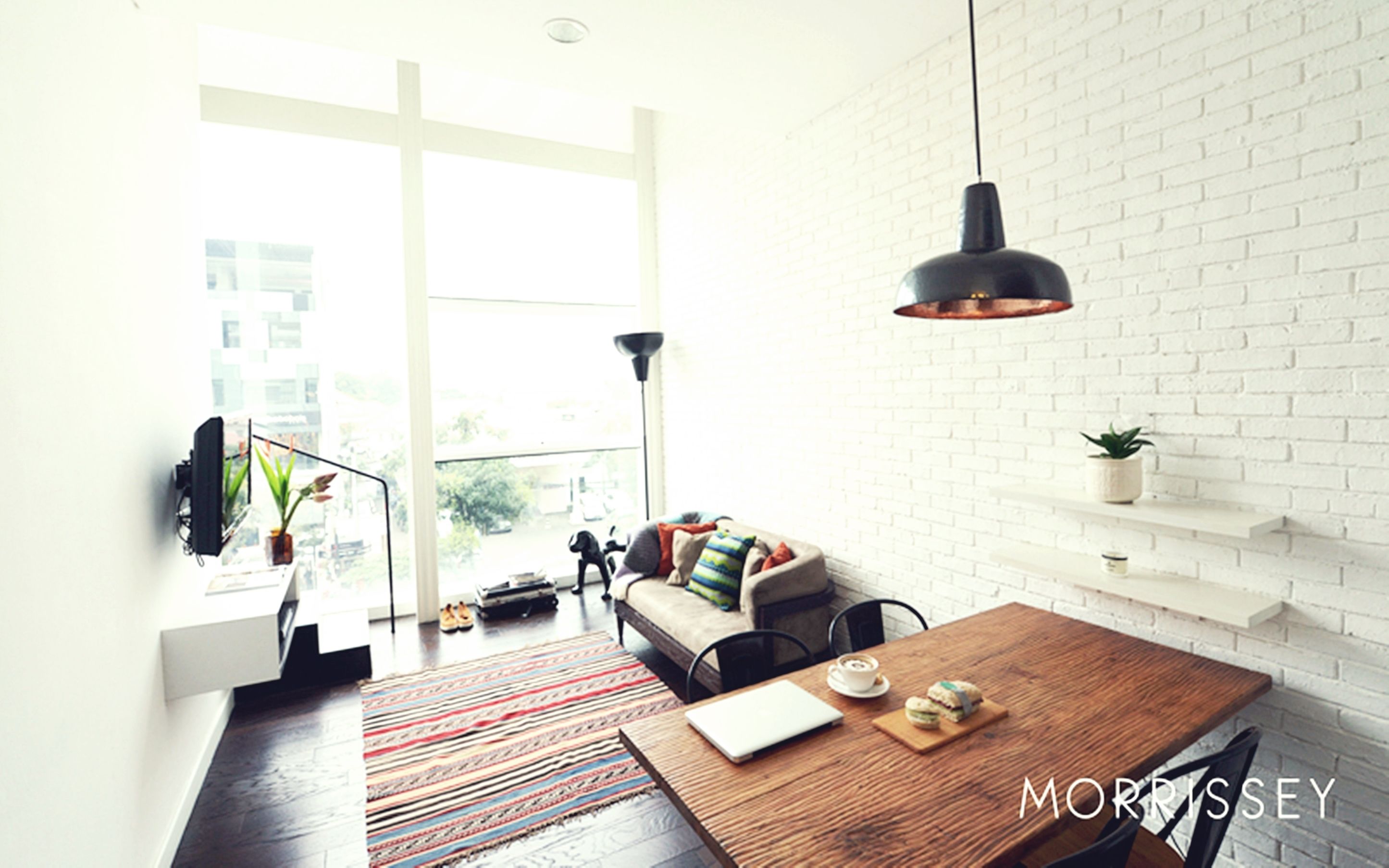 Uma sala de estar moderna com uma mesa de jantar de madeira, um sofá com almofadas coloridas, uma janela grande e uma parede de tijolos brancos.